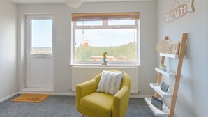 Um quarto com uma poltrona amarela, uma janela com vista para a praia, um radiador branco e uma estante de madeira com objetos.