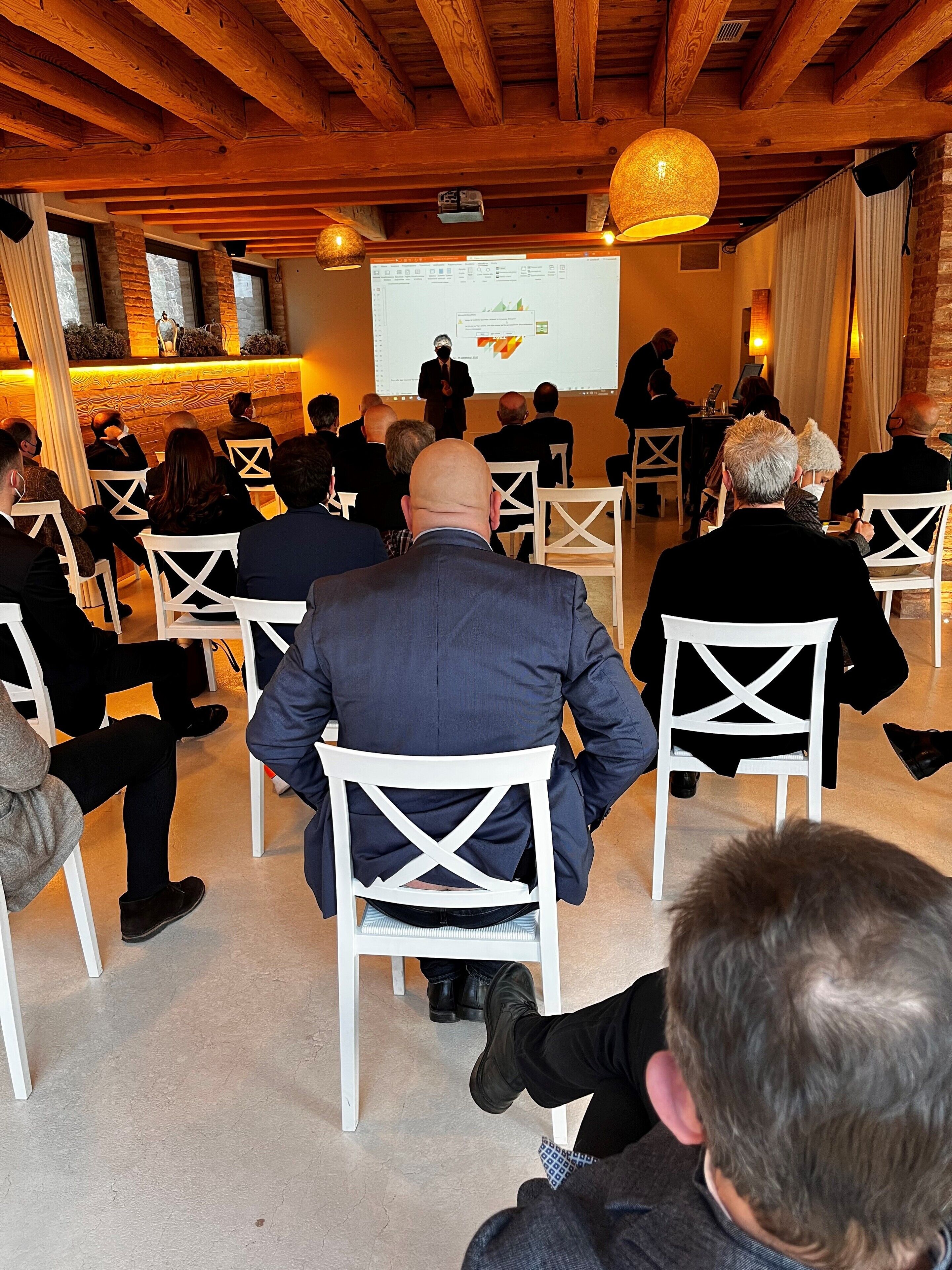 Sala de conferências com participantes sentados em cadeiras brancas, voltados para uma tela de apresentação.