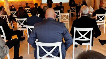 Sala de conferências com participantes sentados em cadeiras brancas, voltados para uma tela de apresentação.
