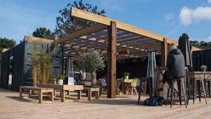 Uma área de pátio ao ar livre com mesas e bancos de madeira, uma estrutura de pérgola e plantas em vasos.