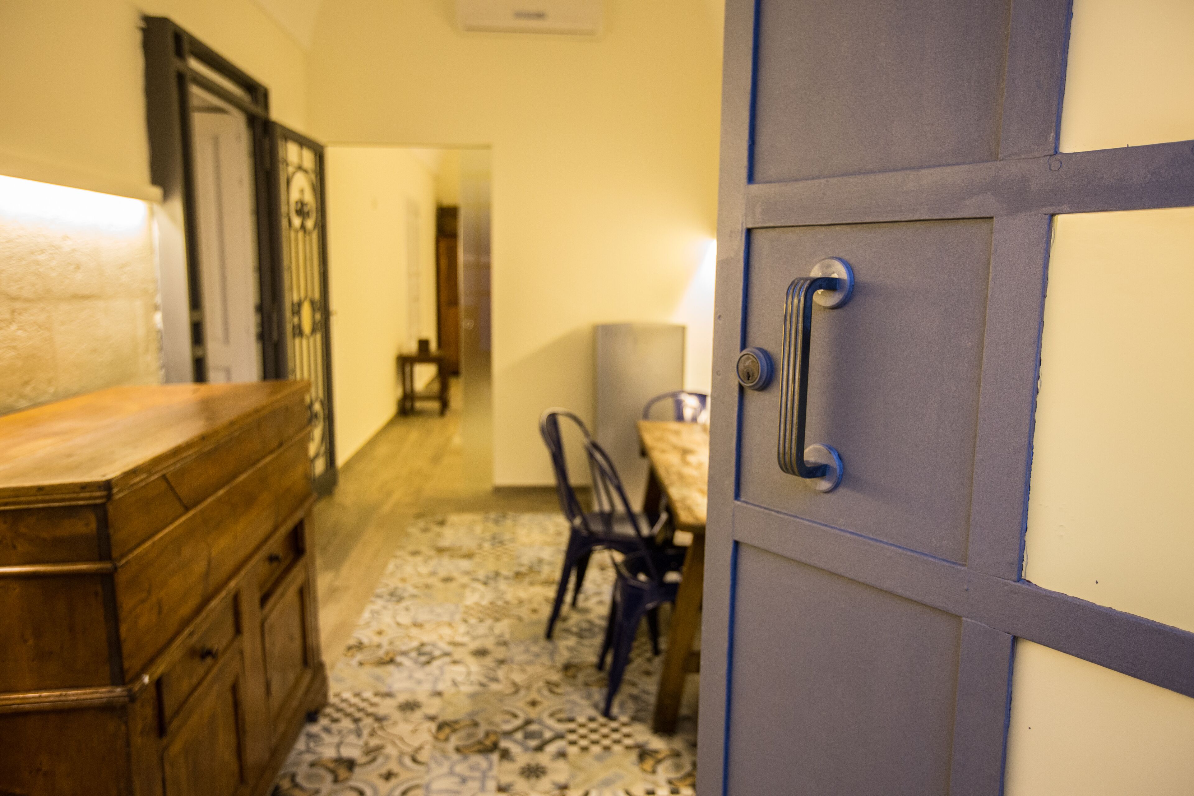 Um corredor com cômoda de madeira, piso estampado e porta azul com puxador.