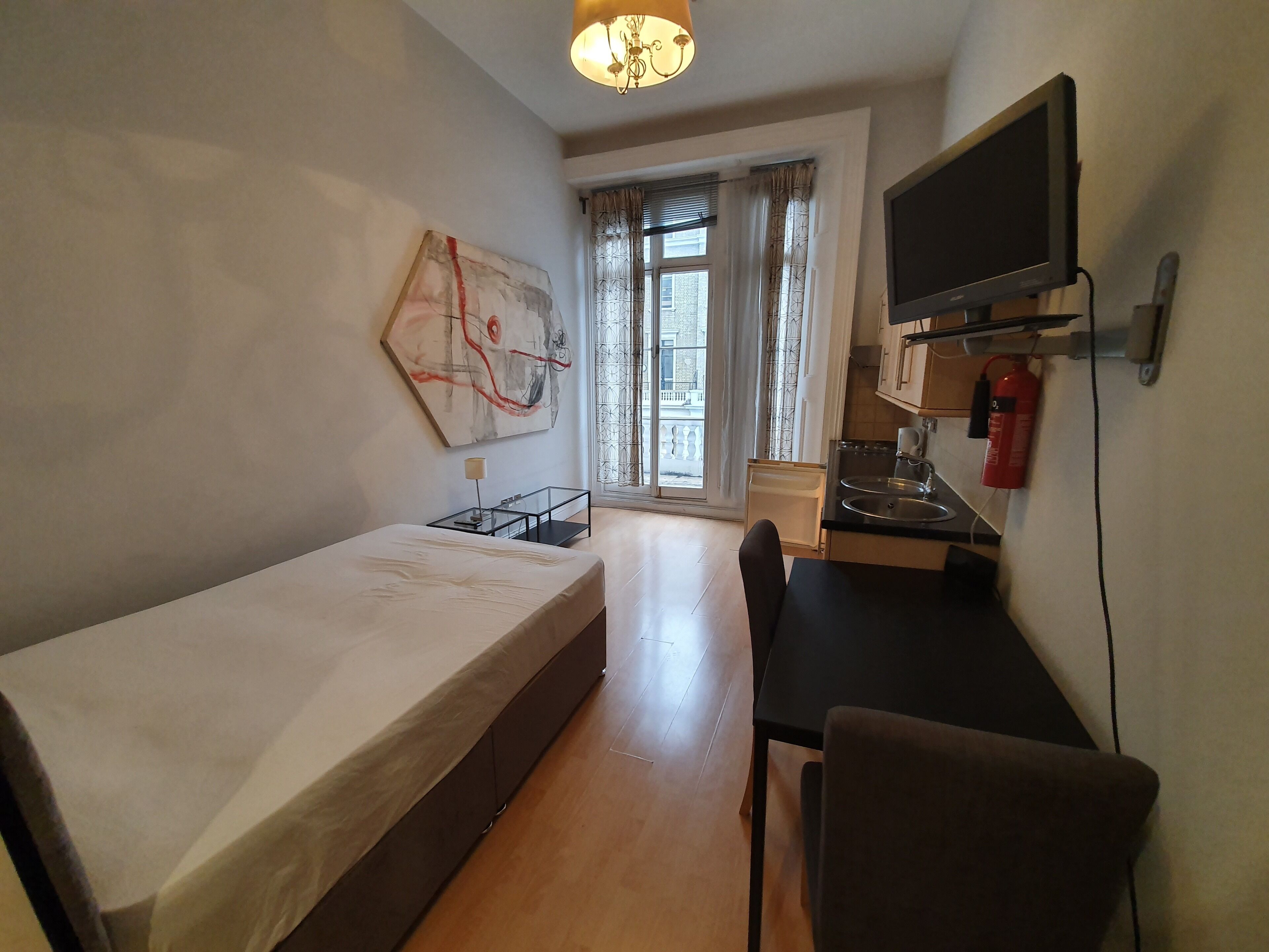 Quarto pequeno e compacto com uma cama, uma escrivaninha, uma cadeira, uma televisão e uma janela com cortinas.