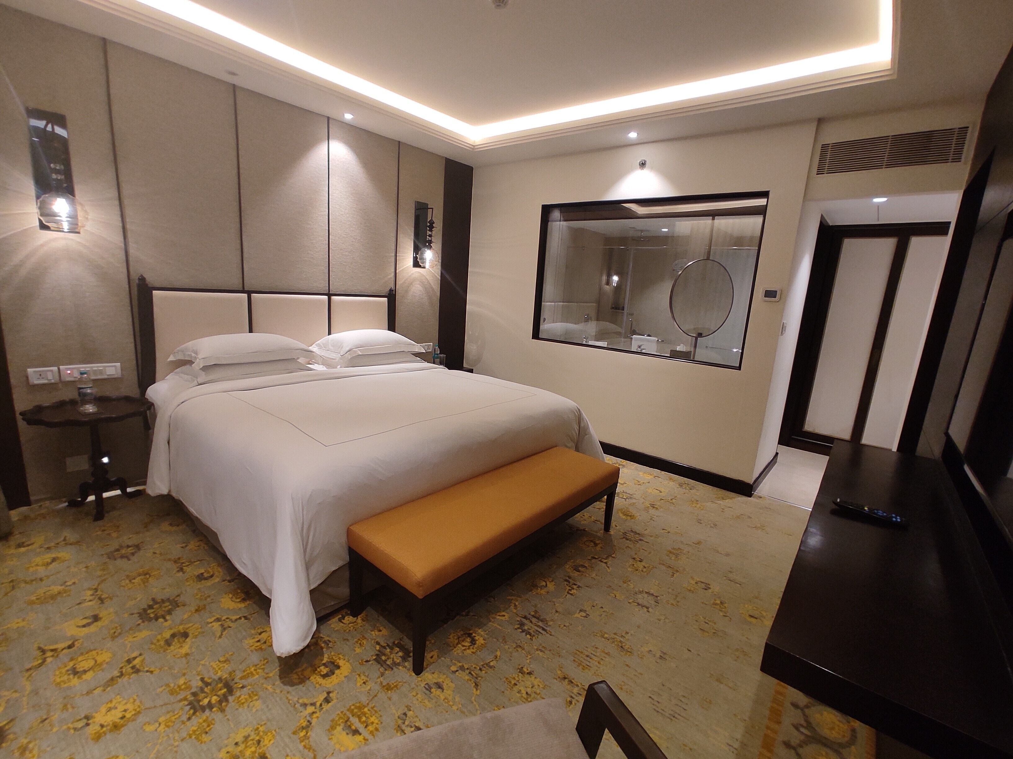 Quarto de hotel com cama grande, mesa de cabeceira, um banco, espelho e televisão.