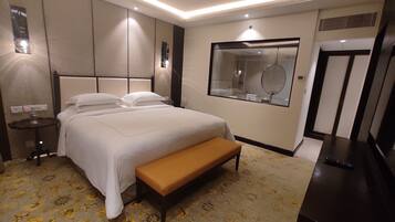 Quarto de hotel com cama grande, mesa de cabeceira, um banco, espelho e televisão.