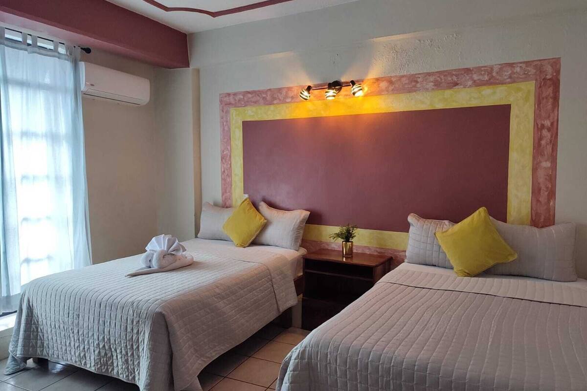 Quarto de hotel com duas camas, uma mesinha de cabeceira e uma janela grande com cortinas.