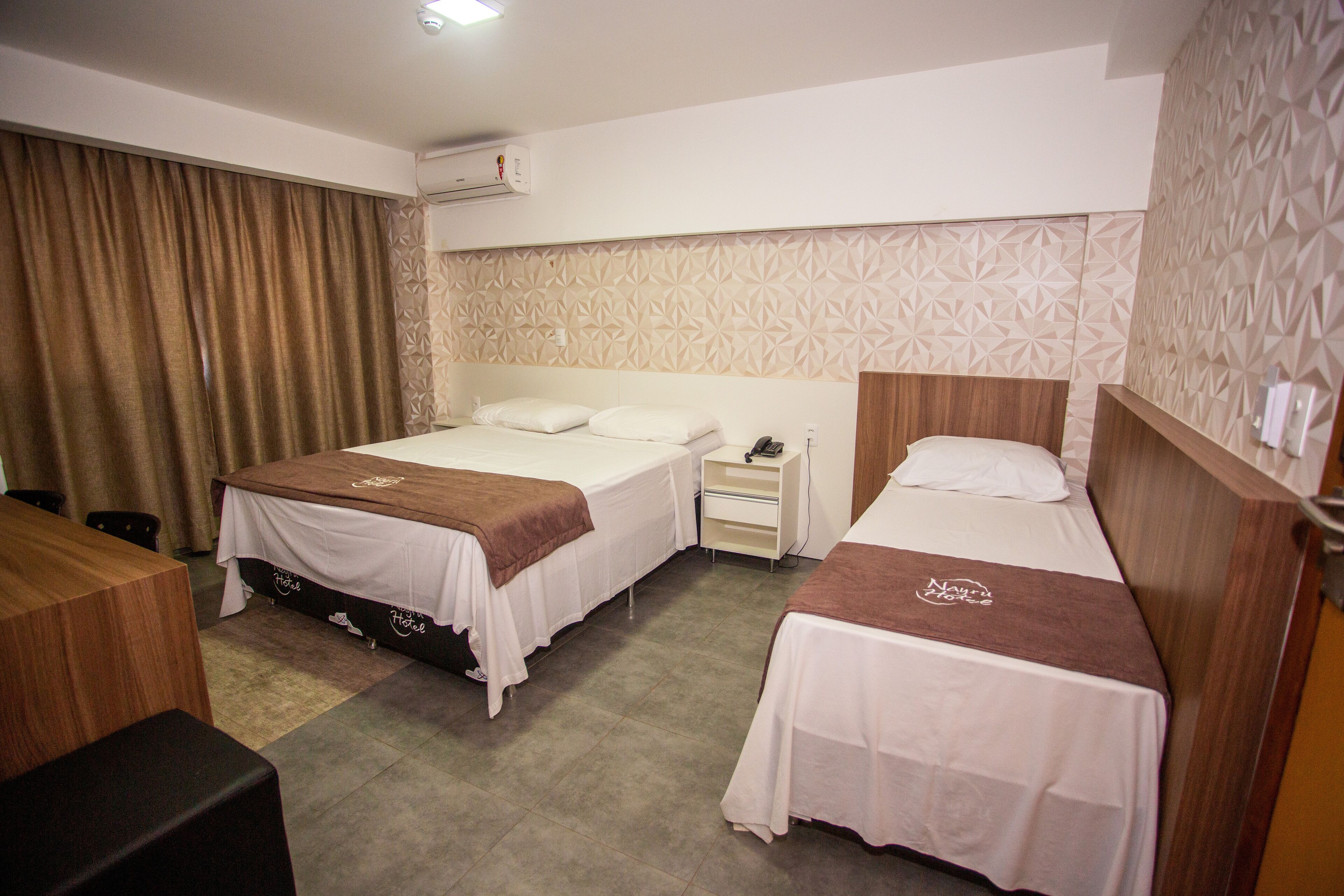 Quarto de hotel com duas camas, uma escrivaninha de madeira e um criado-mudo com telefone.