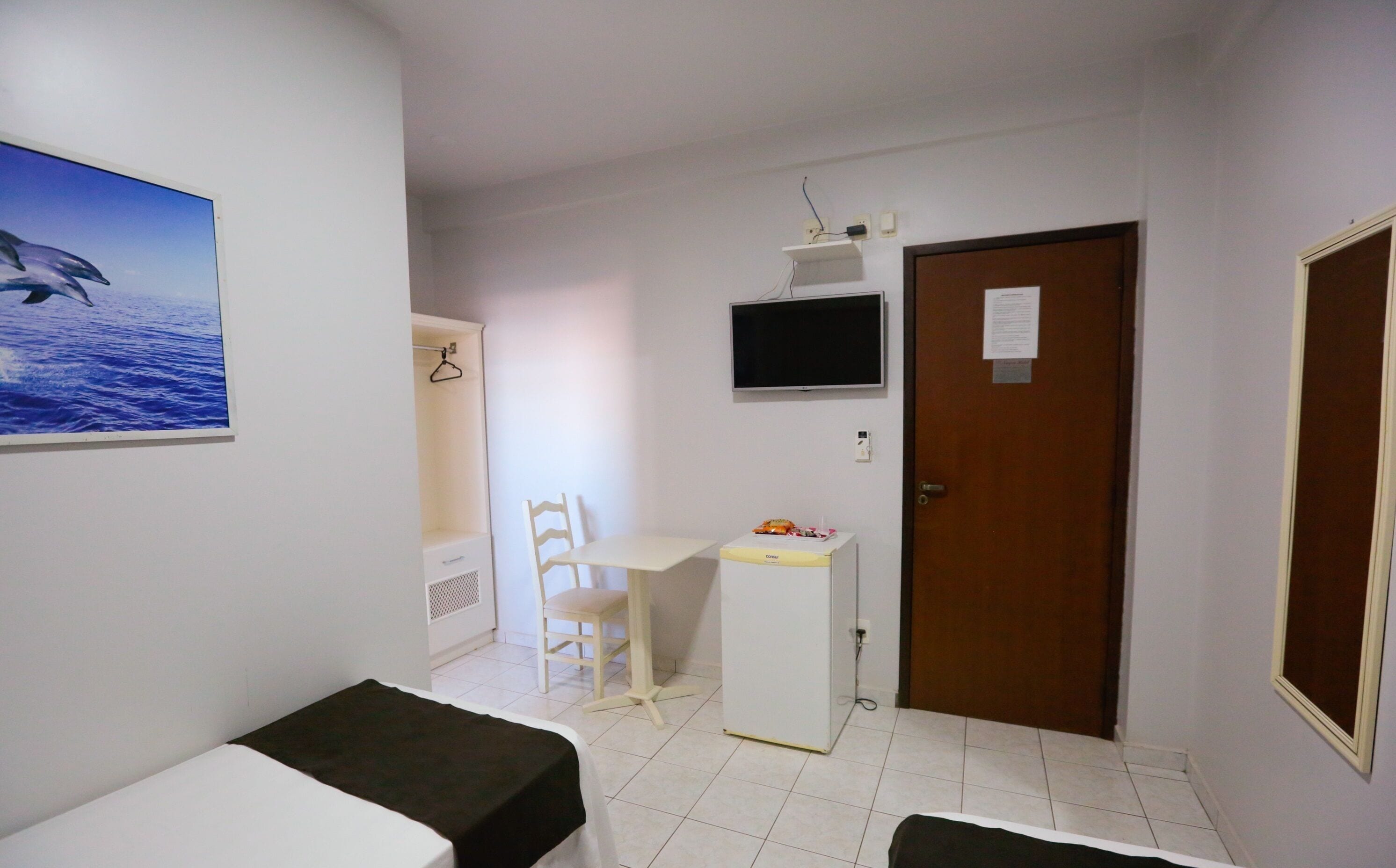 Quarto pequeno e limpo com uma cama, escrivaninha, cadeira, televisão e porta.