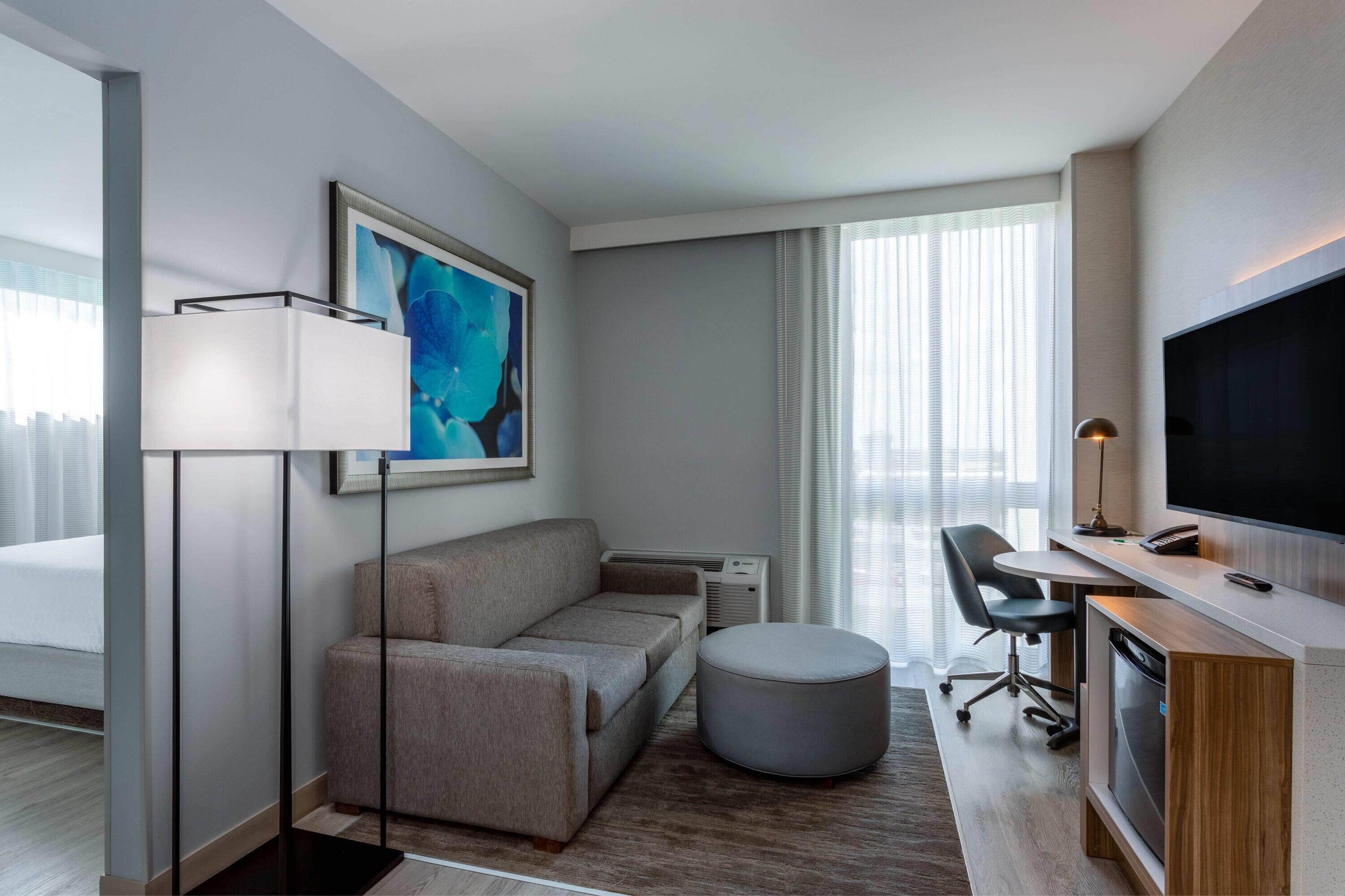 Quarto de hotel moderno com sofá, banquinho, televisão, escrivaninha e ar condicionado.