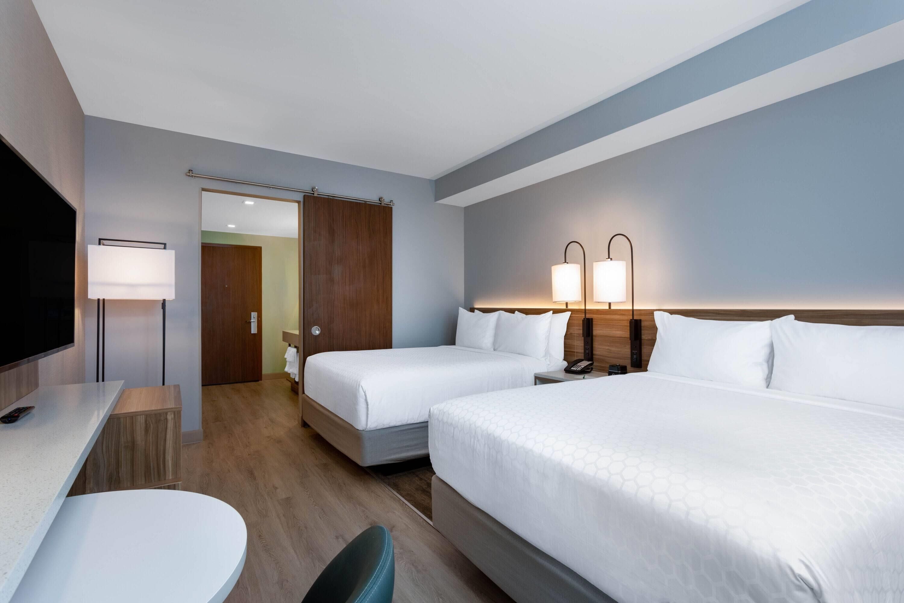 Quarto de hotel com duas camas, uma escrivaninha, uma cadeira, uma televisão e a porta do banheiro.