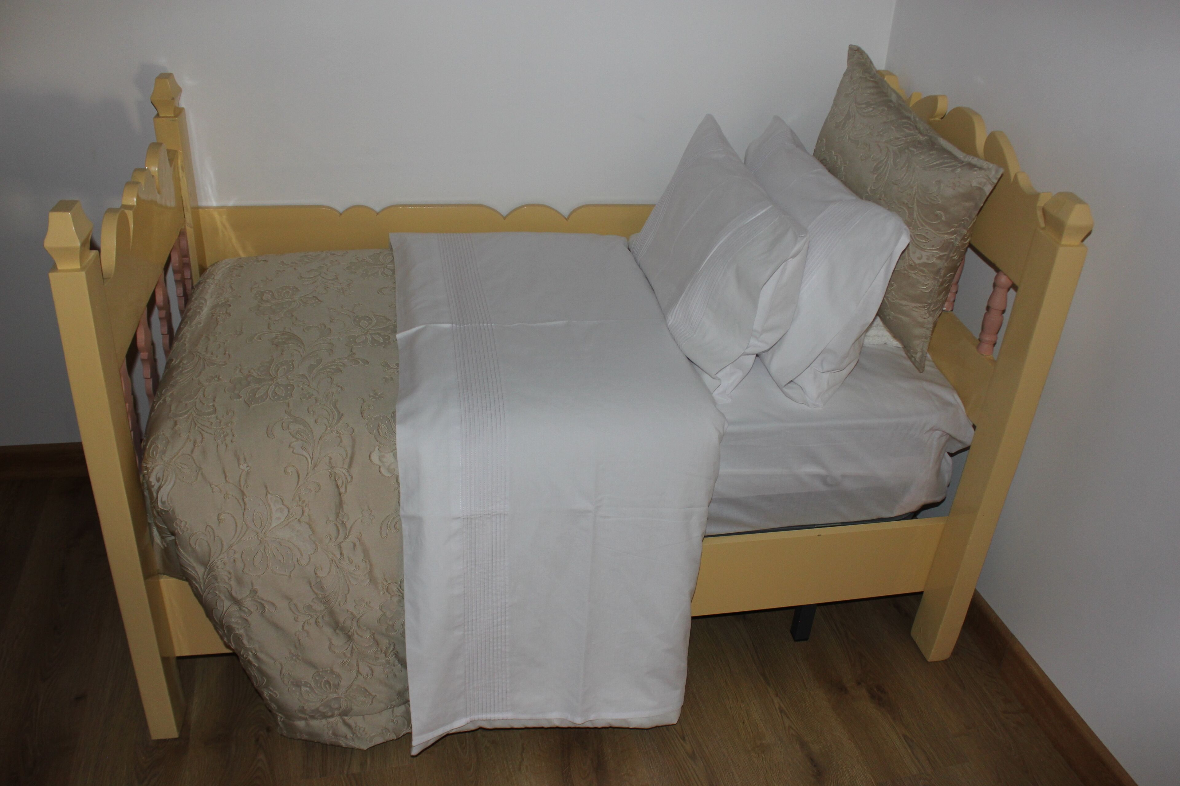 Uma cama de solteiro com estrutura de madeira, colcha bege e duas almofadas.