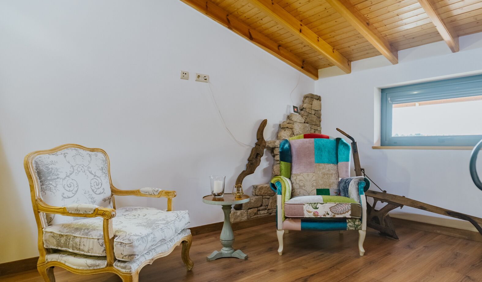 Quarto com teto de madeira, parede de pedra, cadeira rústica, poltrona colorida com acolchoado e uma pequena mesa com uma vela.