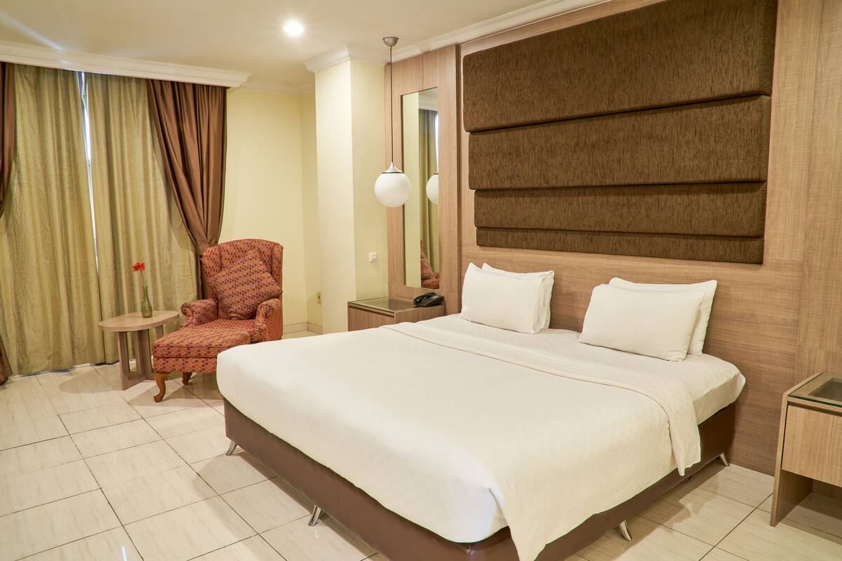 Quarto de hotel com uma cama grande, uma cadeira, uma mesinha e um espelho.