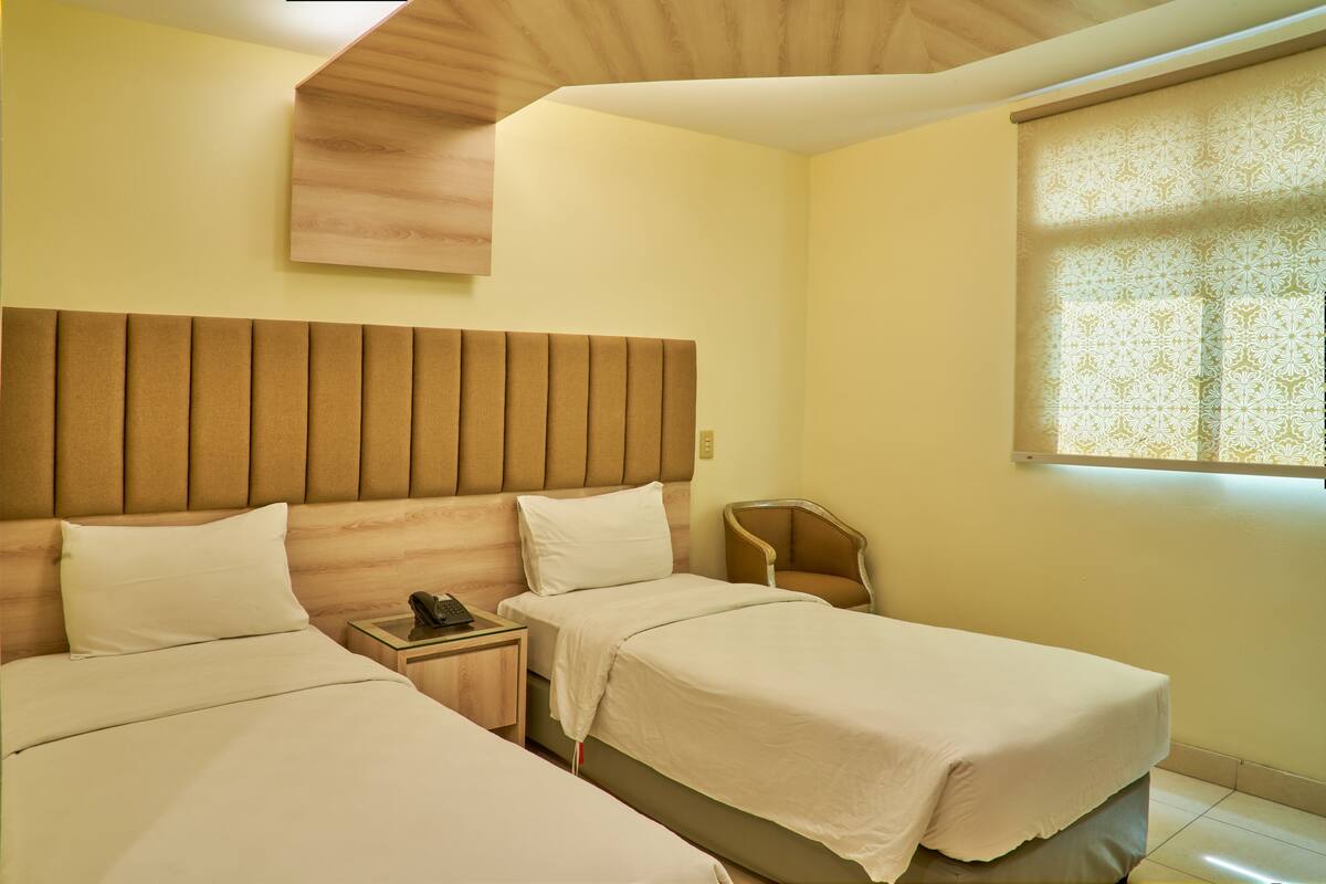 Quarto de hotel com duas camas, cabeceira de madeira, mesa de cabeceira com telefone e janela com cortinas estampadas.