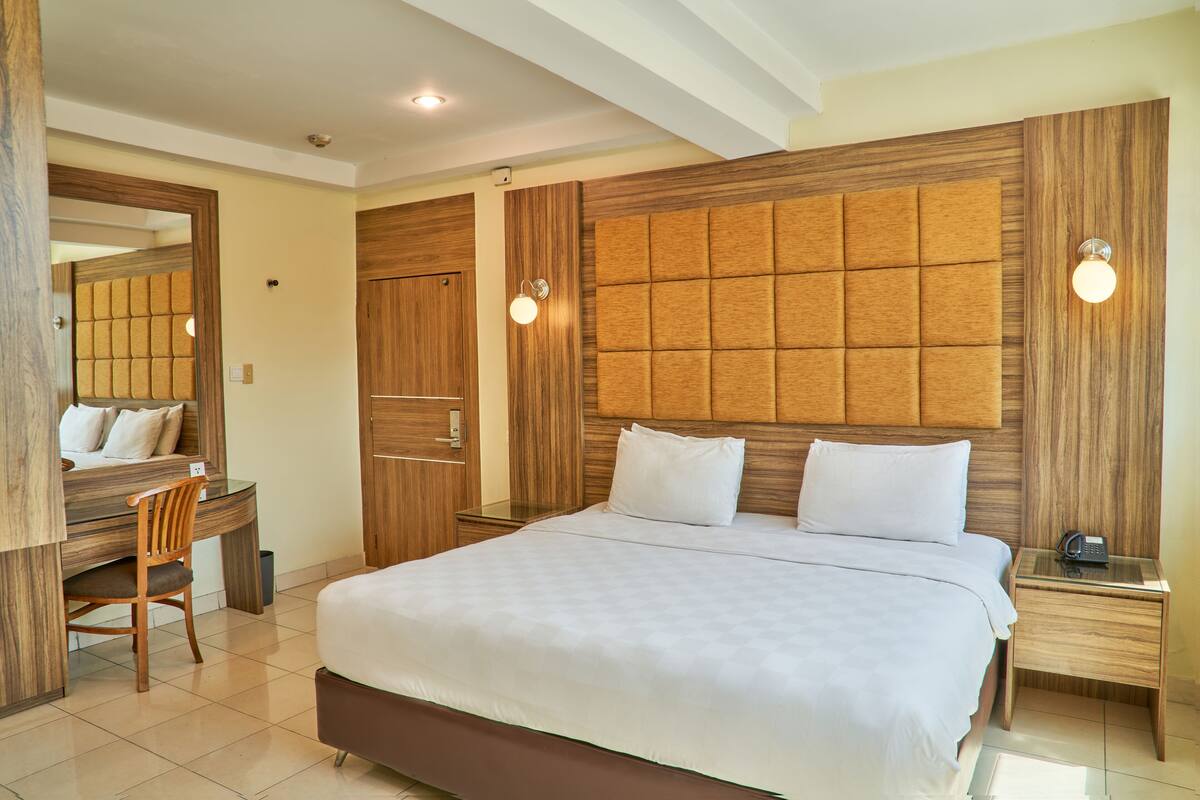 Quarto de hotel com uma cama grande, cabeceira de madeira, escrivaninha com cadeira, mesa de cabeceira com telefone e um espelho.