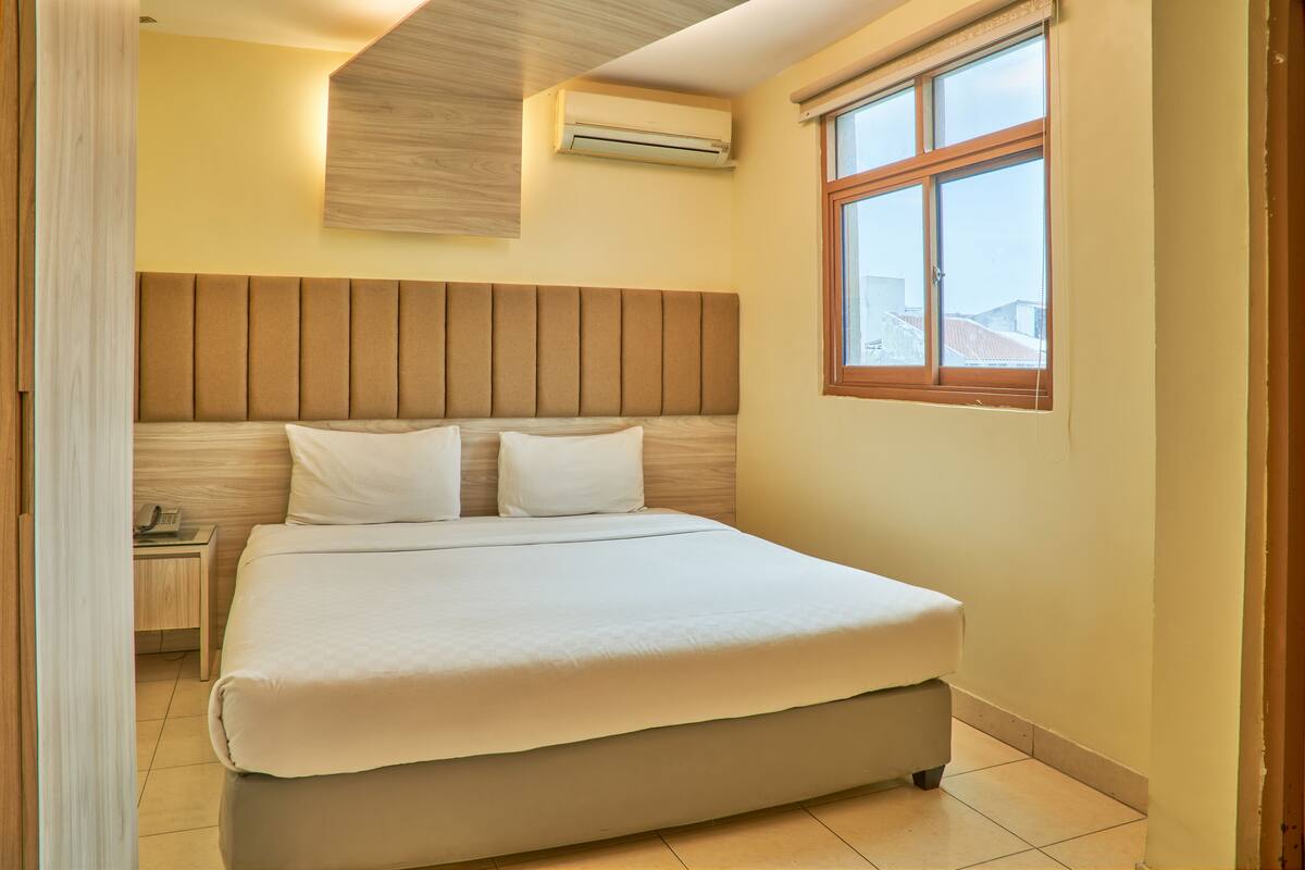 Cama bem arrumada com cabeceira, mesa de cabeceira com telefone e janela com vista para edifícios.