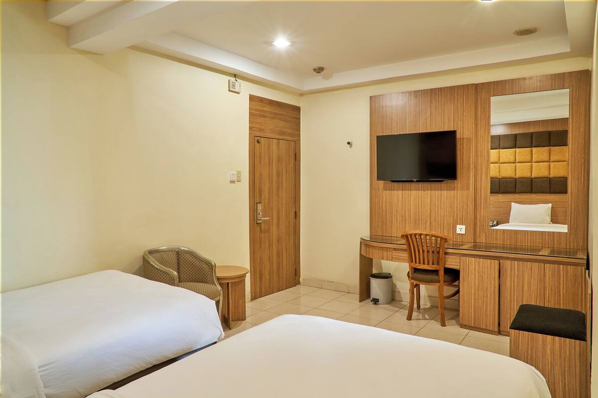Quarto de hotel com duas camas, uma escrivaninha, uma cadeira, uma televisão e um guarda-roupa de madeira.