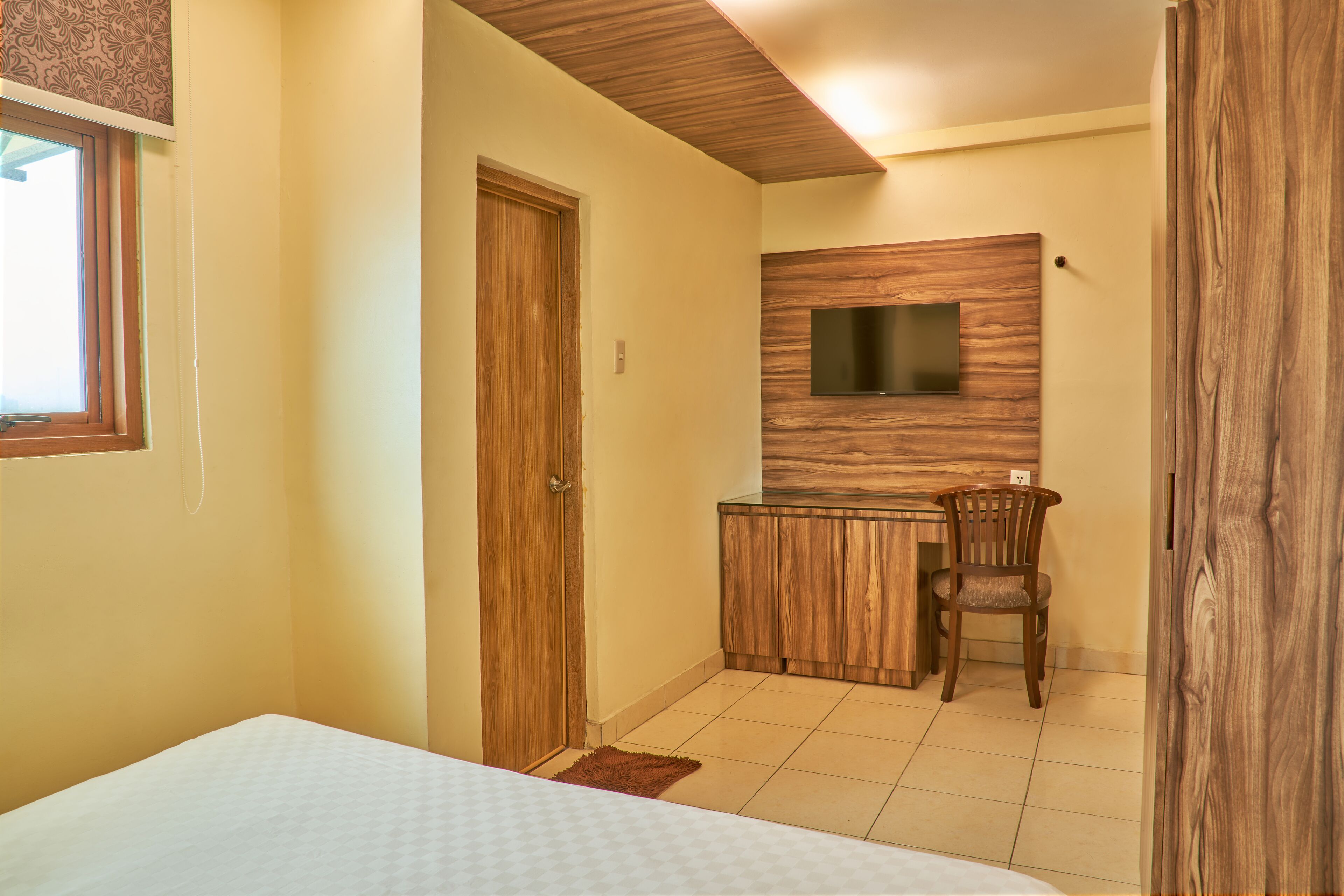 Quarto com uma cama, uma escrivaninha de madeira, uma cadeira e uma televisão na parede.