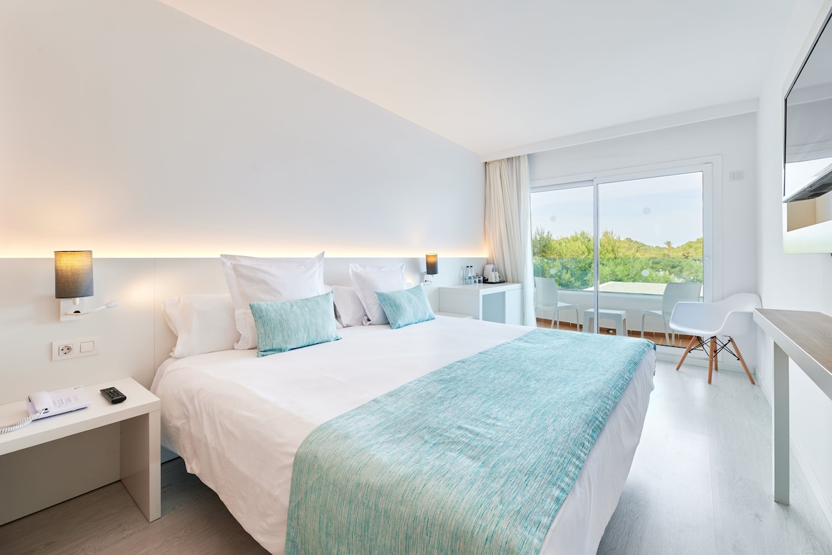 Quarto de hotel moderno com cama grande, mesas de cabeceira, escrivaninha e varanda com vista.