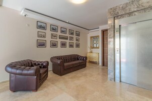 Um hall amplo com um sofá de couro marrom, uma poltrona menor e piso de mármore. Há quadros nas paredes e um elevador à direita.