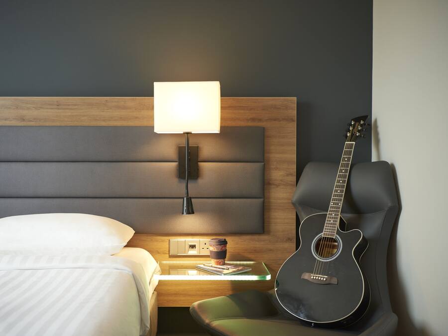 Quarto de hotel com uma cama, uma mesa de cabeceira com abajur, um violão e uma xícara de café.