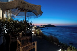 Uma área externa com mesas e cadeiras, um guarda-sol de vime e vista para o oceano ao entardecer.