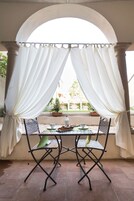 Área de estar coberta ao ar livre com duas cadeiras e uma mesa, oferecendo vista pela janela com cortinas brancas.