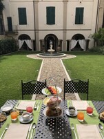 Uma mesa de jantar com um arranjo de frutas e doces em três níveis, uma fonte ao fundo e um gramado bem cuidado.