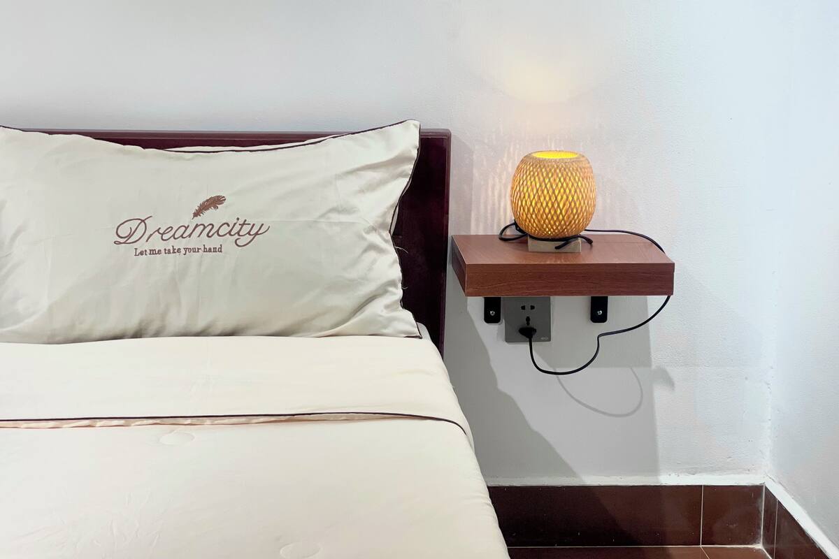 Uma cama com travesseiro que diz "Dreamcity Let me take your hand" e uma mesa de cabeceira com luminária.