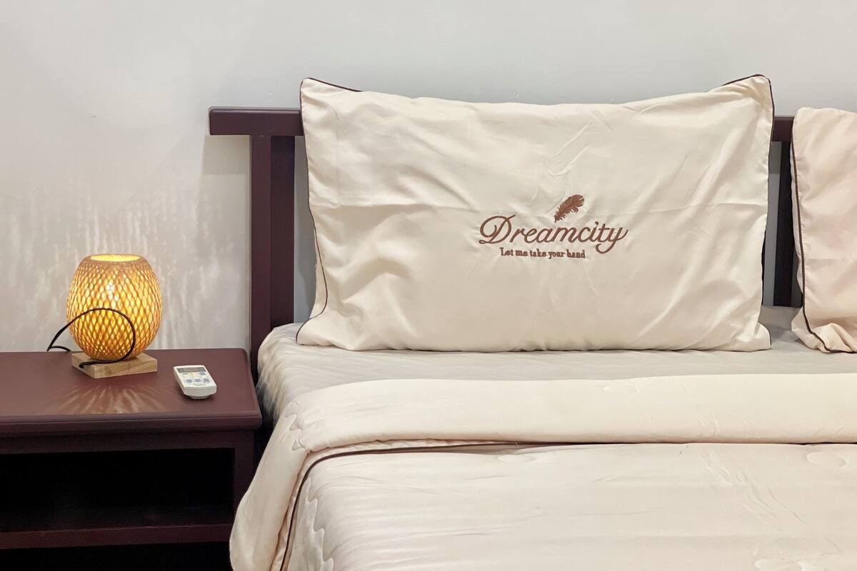 Uma cama bem arrumada com um travesseiro exibindo o texto "Dreamcity Let me take your hand". Uma mesa de cabeceira acomoda uma luminária e um controle remoto.