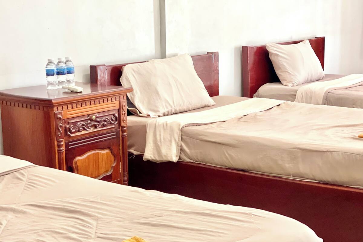 Quarto de hotel com duas camas, uma cômoda de madeira e uma mesinha de cabeceira com garrafas de água.
