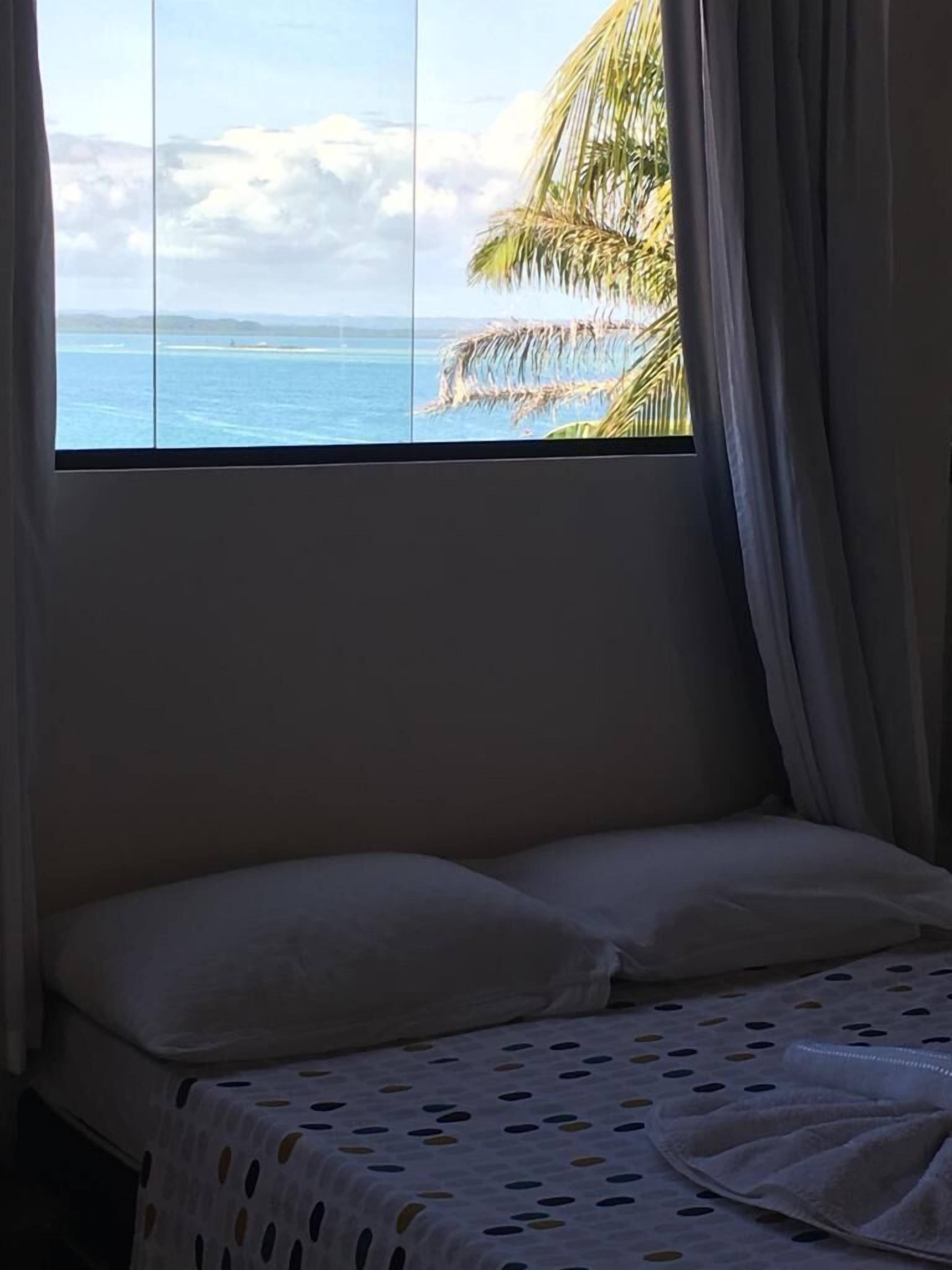 Quarto com cama, janela com cortinas e vista para o oceano.