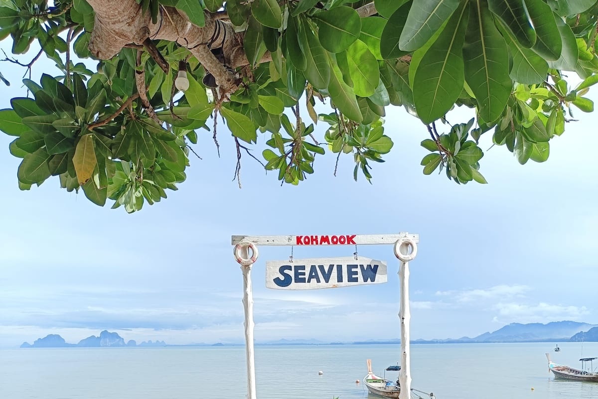Uma placa de madeira com a inscrição "Seaview" e "Kohmook", com vista para um mar calmo, barcos e uma praia ao fundo.