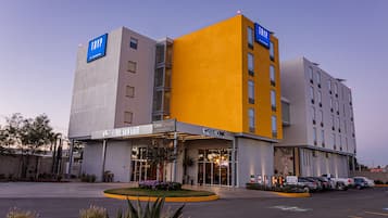 Um hotel moderno com fachada marcante em amarelo e cinza, estacionamento e área de entrada ajardinada.