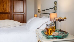 Uma cama bem arrumada com lençóis brancos, um abajur de estilo antigo e uma bandeja com potes de mel e petiscos sobre uma mesa rústica.