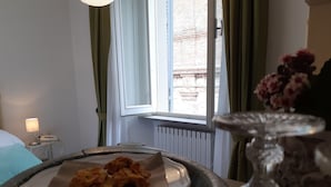 Um quarto com cama, mesa de cabeceira com abajur, janela com cortinas e bandeja com comida.