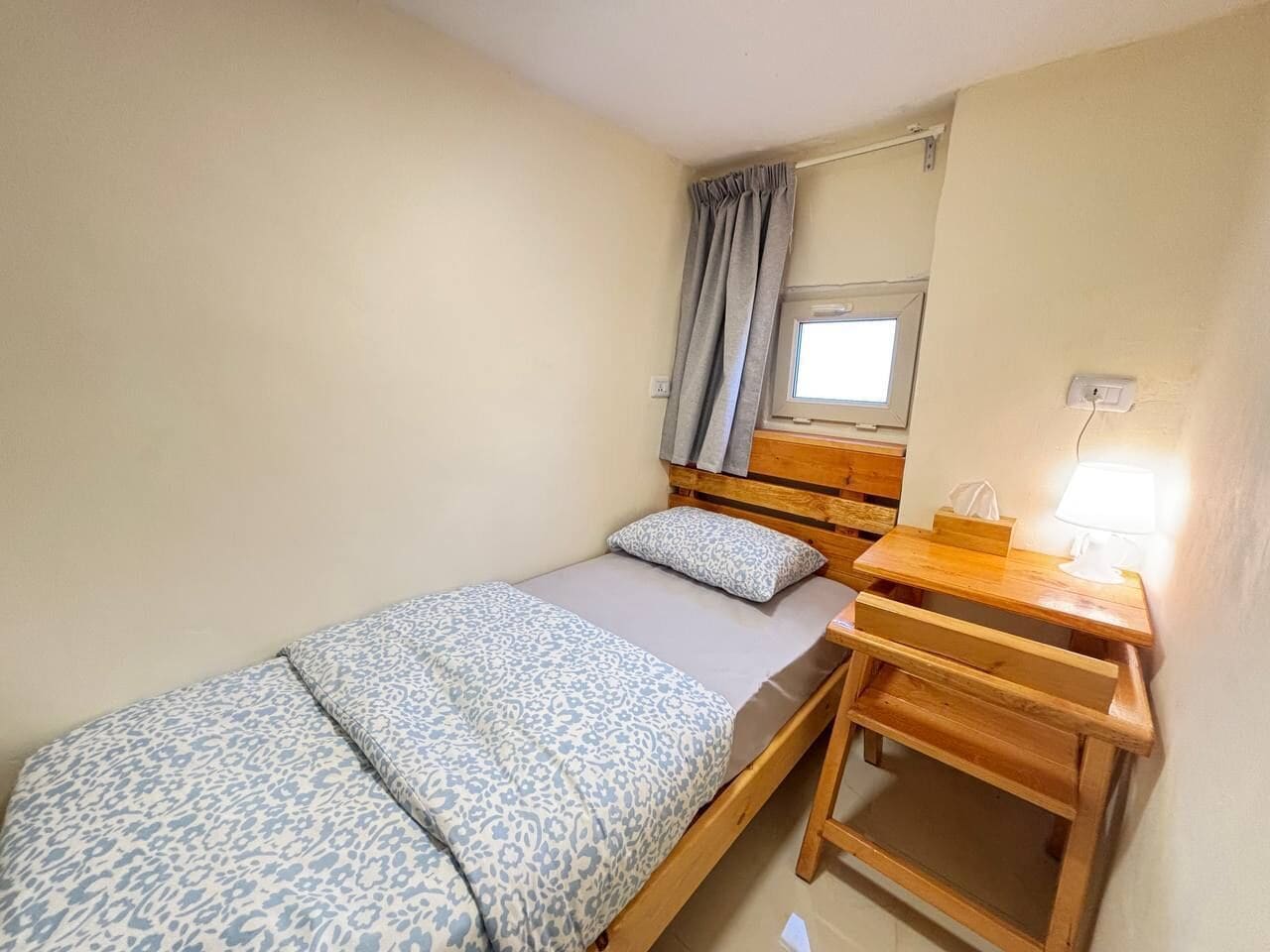Uma cama de solteiro com colcha estampada, uma mesinha de cabeceira com abajur e uma janela com cortinas.