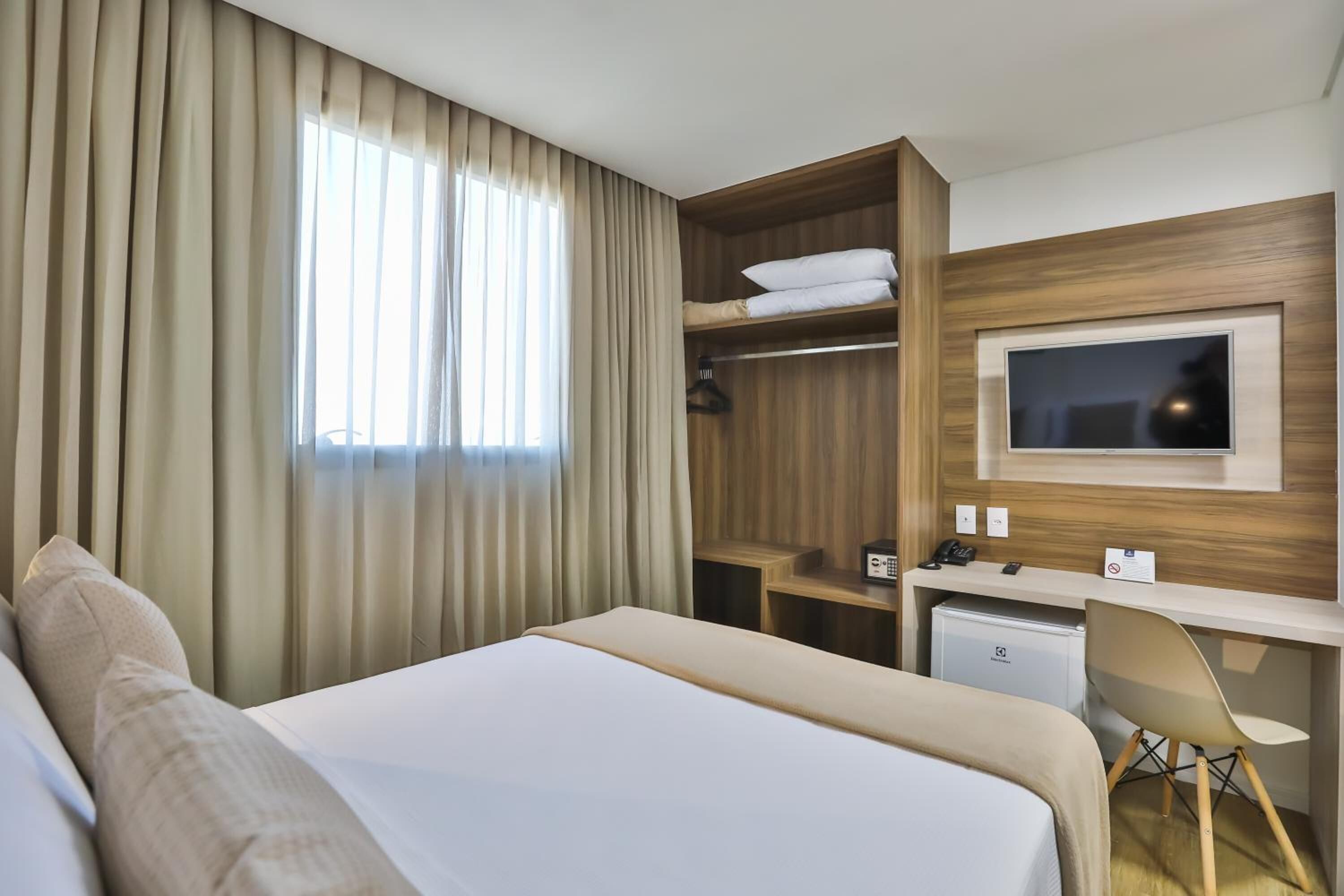 Quarto de hotel com cama, escrivaninha, cadeira, televisão e guarda-roupa.