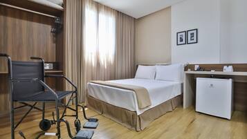 Quarto de hotel com cama, cadeira de rodas, pequeno frigorífico e secretária com cadeira.