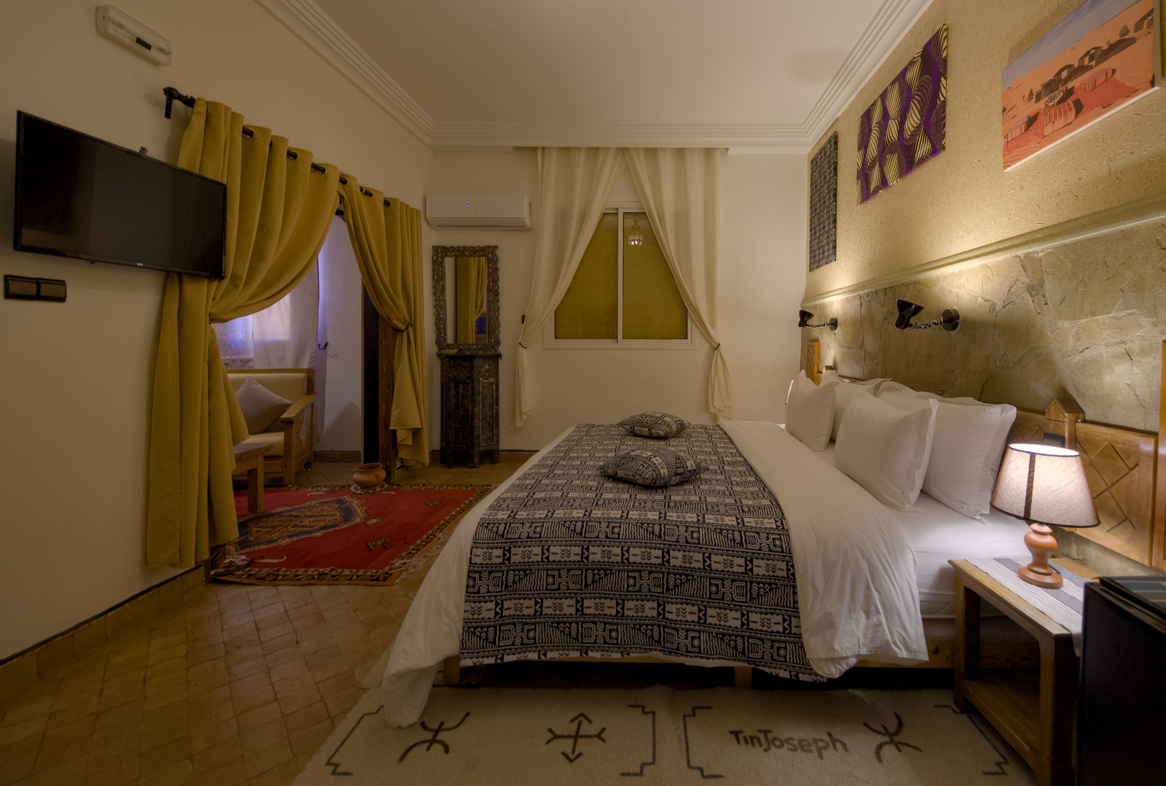 Quarto de hotel com uma cama, televisão, luminária e janela com cortinas.