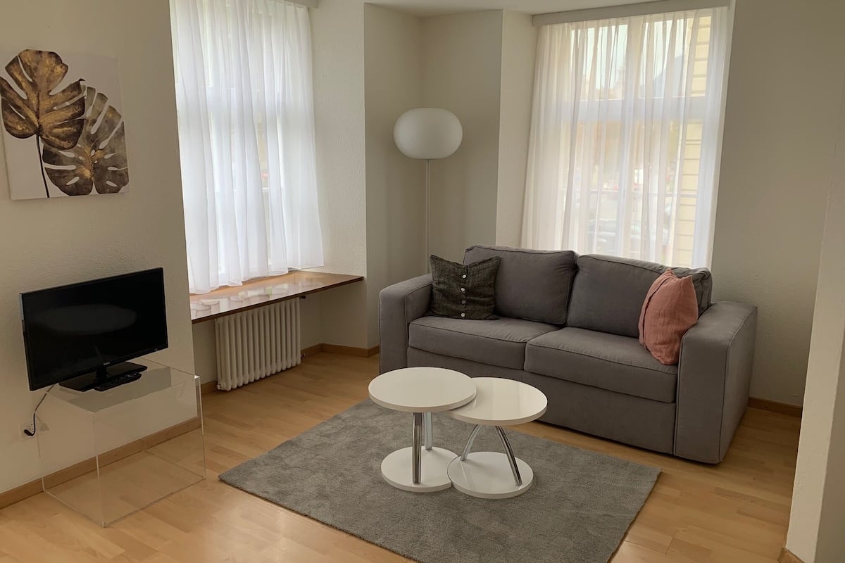 Uma sala de estar moderna com um sofá cinzento, uma mesa redonda branca, uma televisão de tela plana em um suporte de vidro e um radiador.