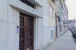 Uma rua de paralelepípedos estreita, ladeada por edifícios, um portão de metal e uma placa.