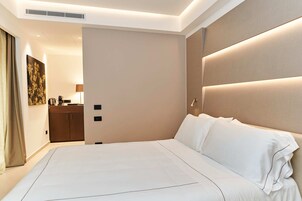 Quarto de hotel moderno com uma cama grande, uma mesinha de cabeceira, uma luminária e vista para o corredor.