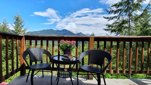 Varanda com duas cadeiras de vime e uma mesa pequena, com vista para uma paisagem montanhosa.