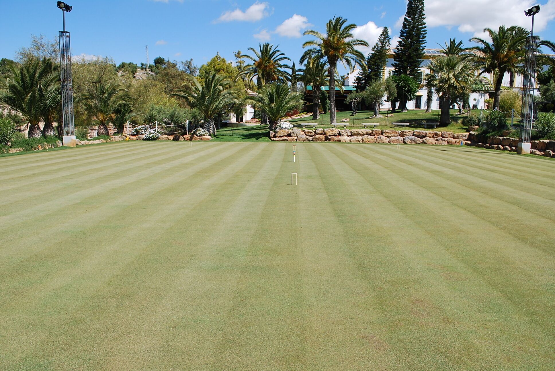 Um campo de golfe bem cuidado com um caminho bem definido, palmeiras e um muro de pedra ao fundo.