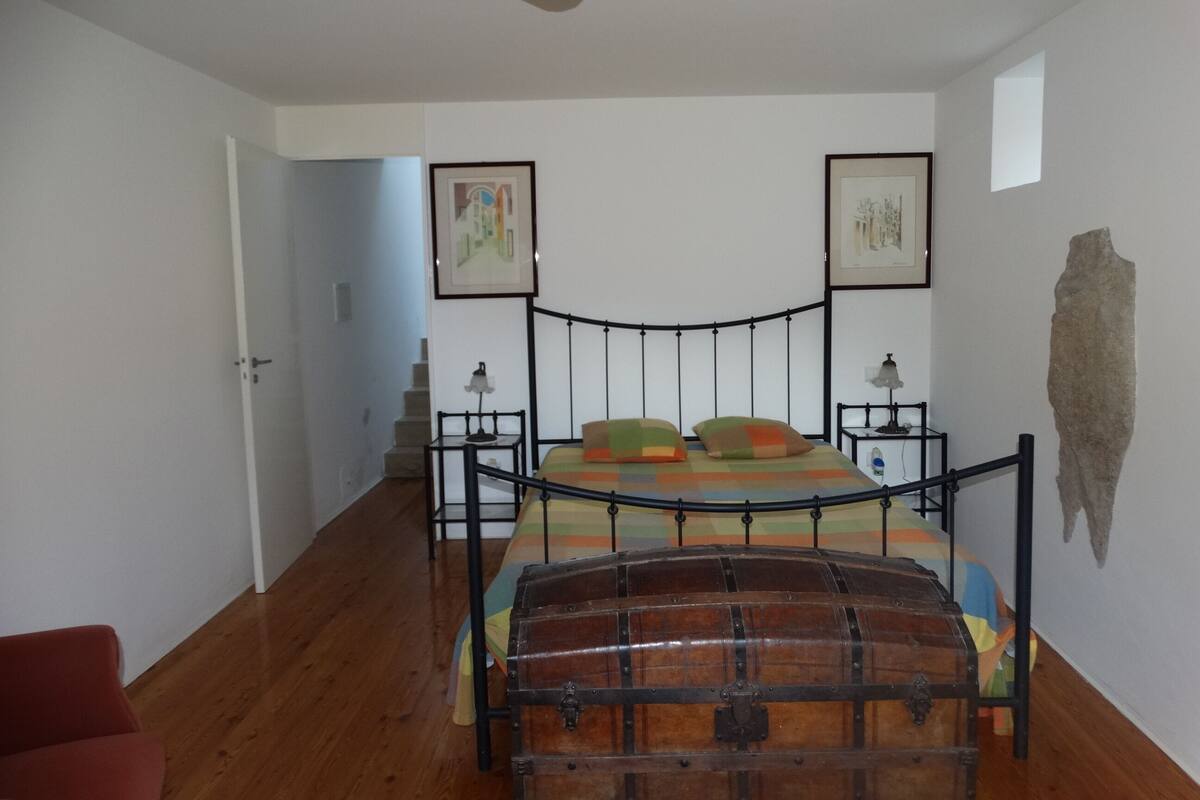 Quarto com uma cama, cabeceira de madeira, duas mesas de cabeceira, uma cadeira vermelha e uma porta que dá para outro cômodo.