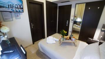 Quarto de hotel com uma cama, uma escrivaninha com controle remoto, um tabuleiro com itens de café da manhã e um espelho grande refletindo o quarto.