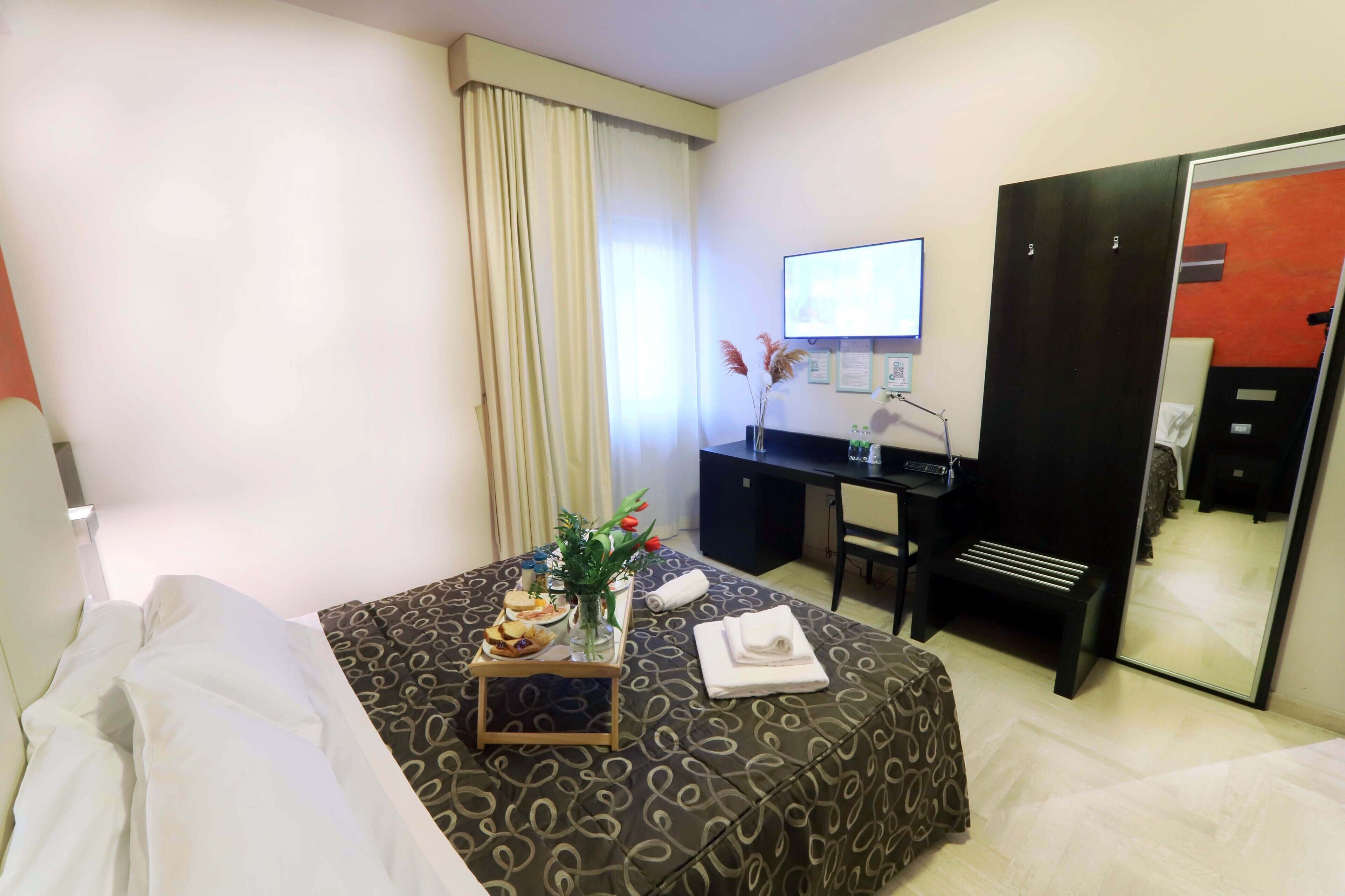 Quarto de hotel com cama, escrivaninha, cadeira, espelho e uma pequena mesa com bandeja de comida.