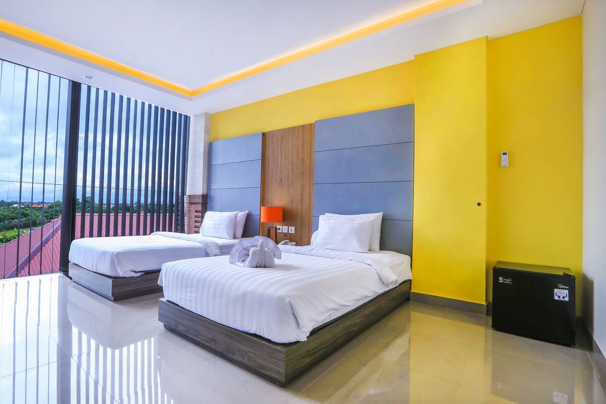 Quarto de hotel moderno com duas camas, uma janela grande com vista e uma parede de destaque amarelo vibrante.
