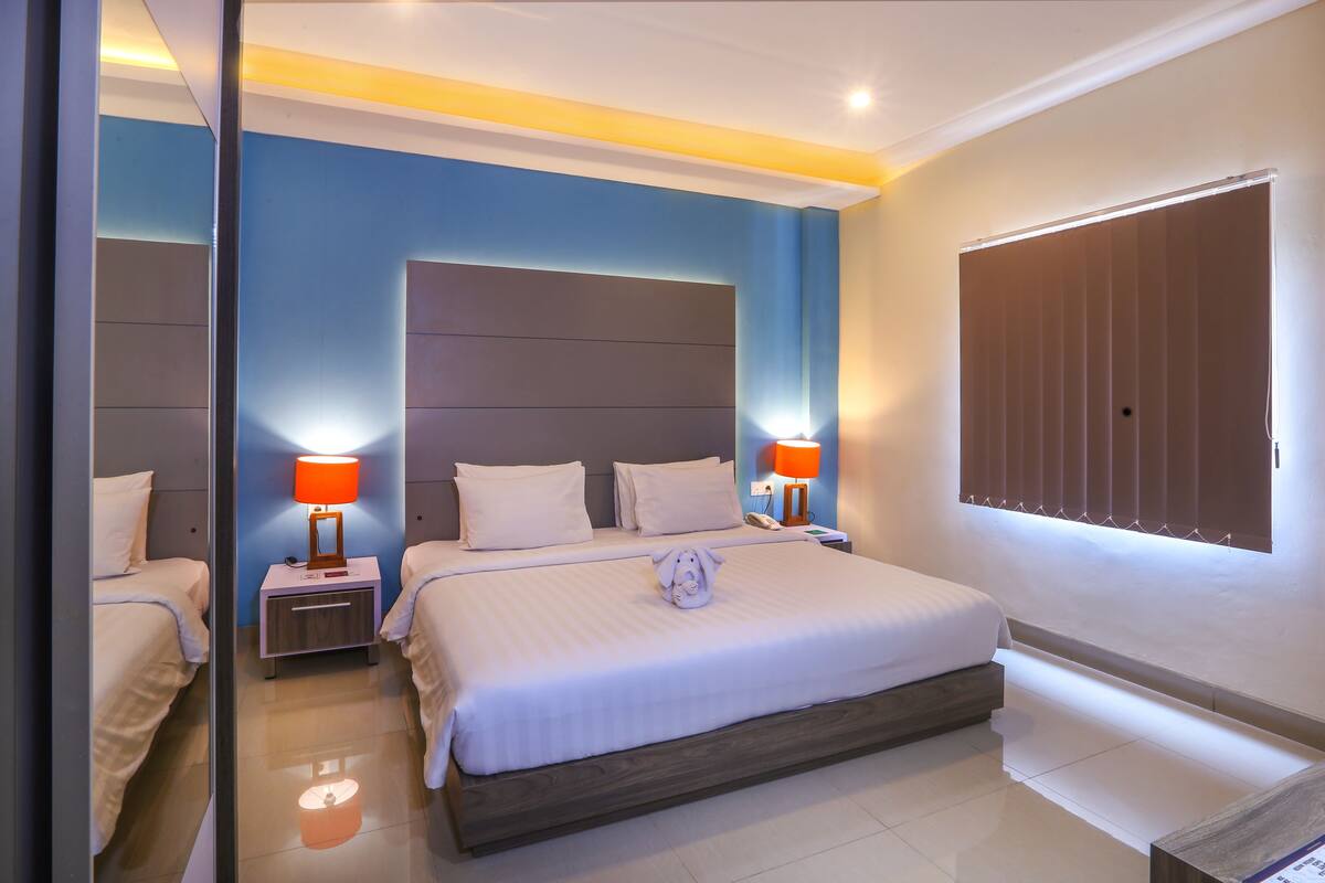 Quarto de hotel com cama grande, mesas de cabeceira com abajures, espelho e janela com persianas.