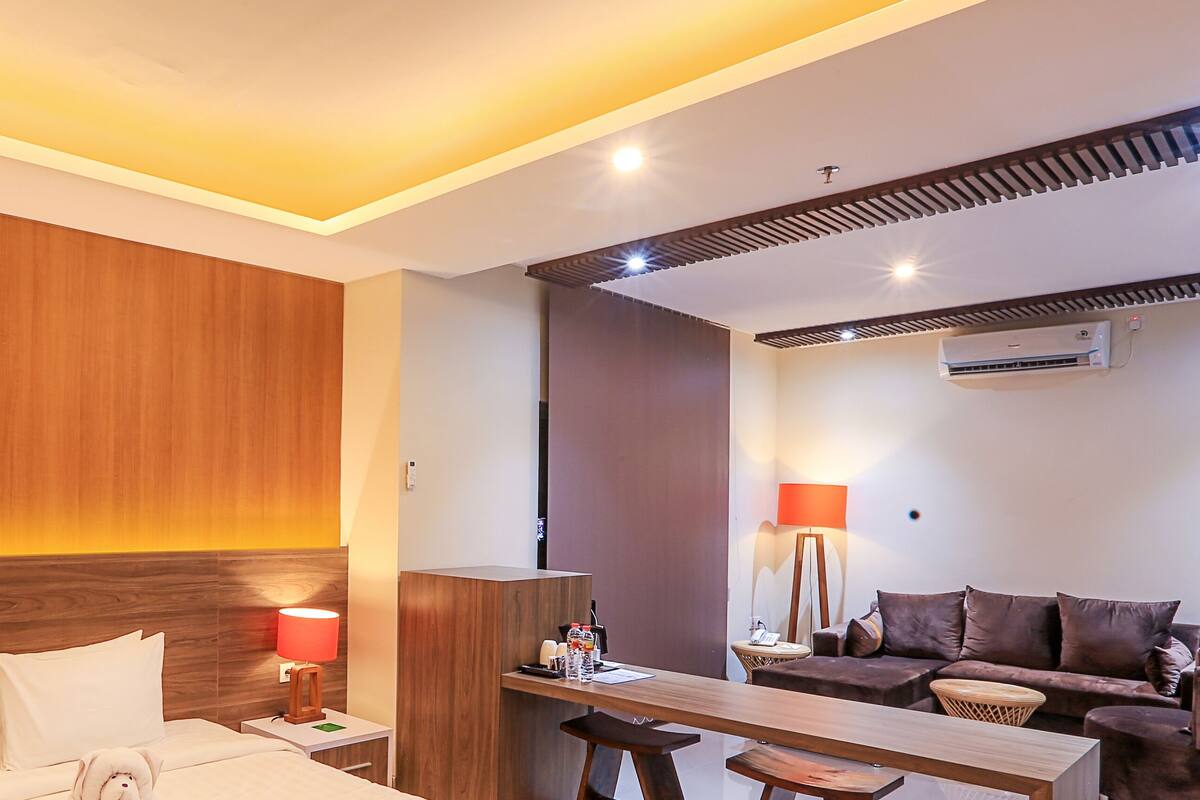 Quarto de hotel com cabeceira de madeira, cama com lençóis brancos, escrivaninha com abajur e sofá.